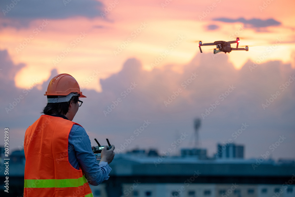 Young Asian engineer flying drone over construction site during sunset ...