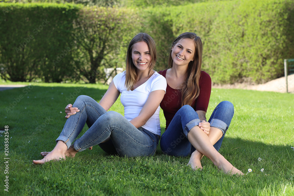 Zwei hübsche junge Frauen sitzen barfuß auf einer Wiese und lachen ...