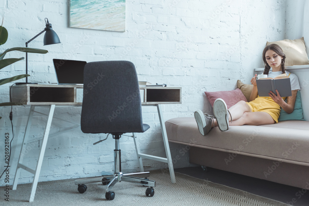 teen girl with cup of coffee reading book on fosa in room with table ...