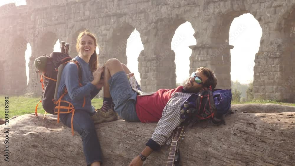 Happy young couple backpackers tourists sitting lying on a log trunk ...