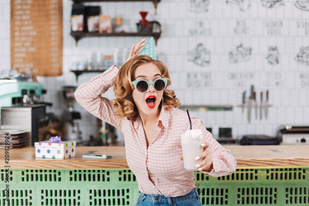 Young surprised lady in sunglasses and birthday cap sitting at the bar ...