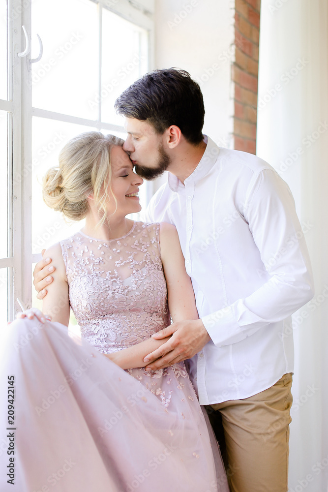 Blonde tender bride wearing dress and standing near window and kissing ...