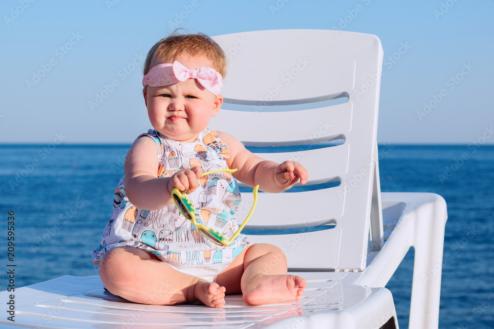 Little baby girl with sunglasses sitting on beach lounge at a sea shore ...