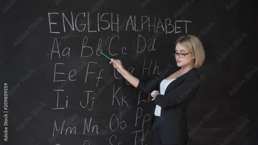 Teacher is writing letter of alphabet on blackboard with chalk ...