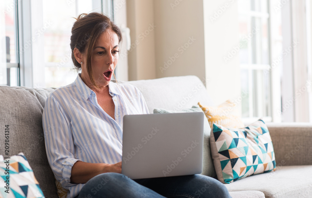 Middle aged woman using laptop in the sofa scared in shock with a ...