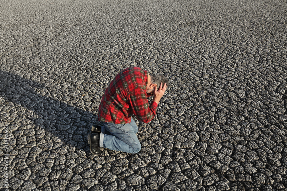 Desperate man kneeling and holding his head in his hands at dry cracked ...
