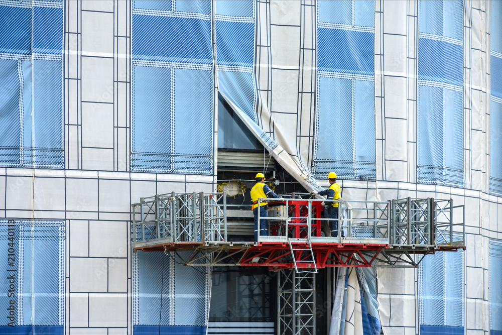 Builders on a high-altitude lift work on the wall of a large building ...