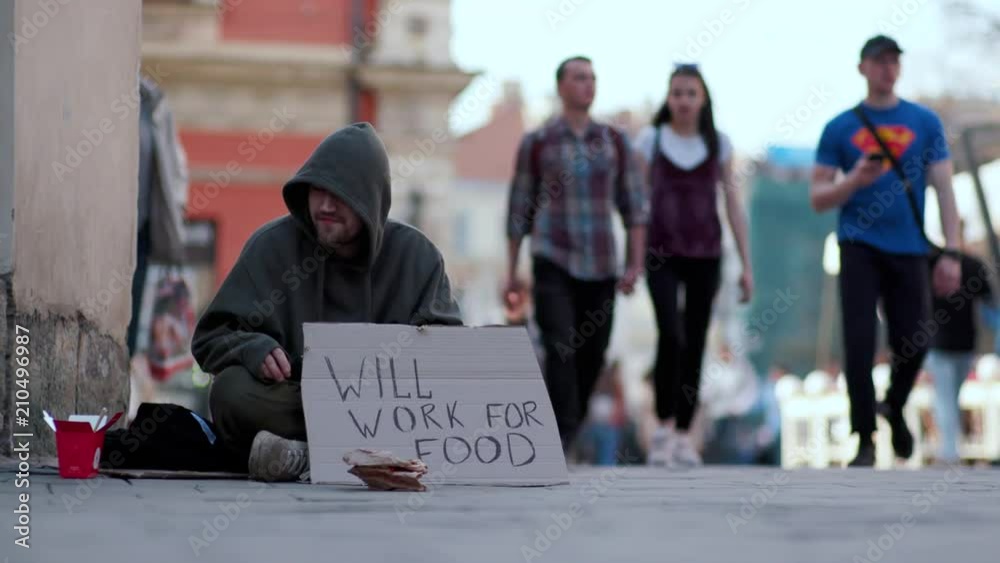 Homeless man sitting on a street with sign eating people walk around ...