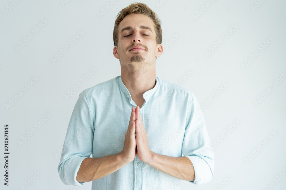 Peaceful young man praying with his eyes closed and keeping hands ...