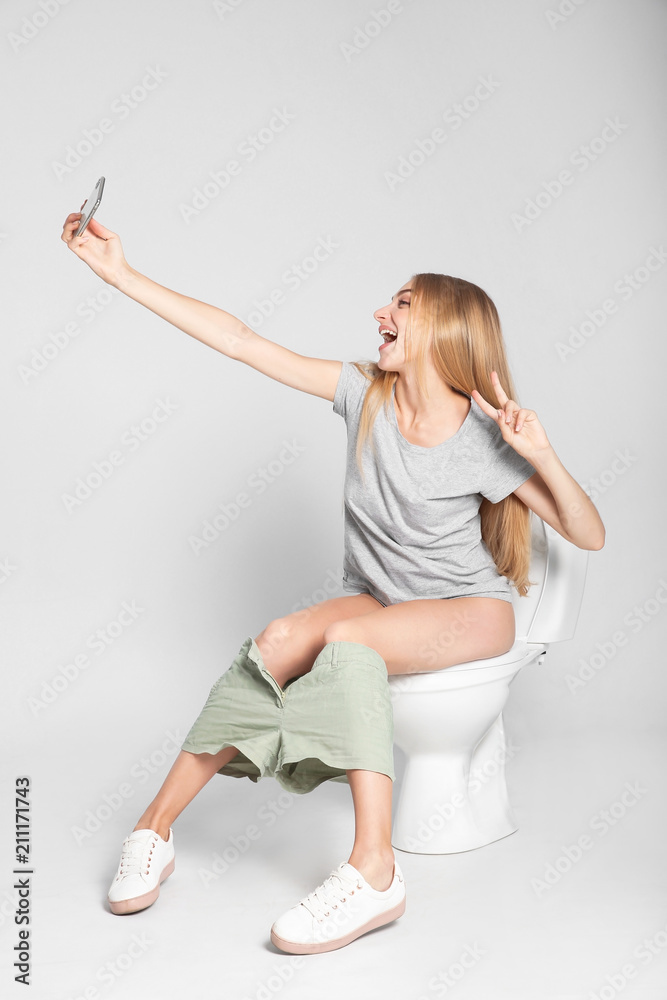 Young woman taking selfie while sitting on toilet bowl against gray ... Young woman taking selfie while sitting on toilet bowl against gray ...