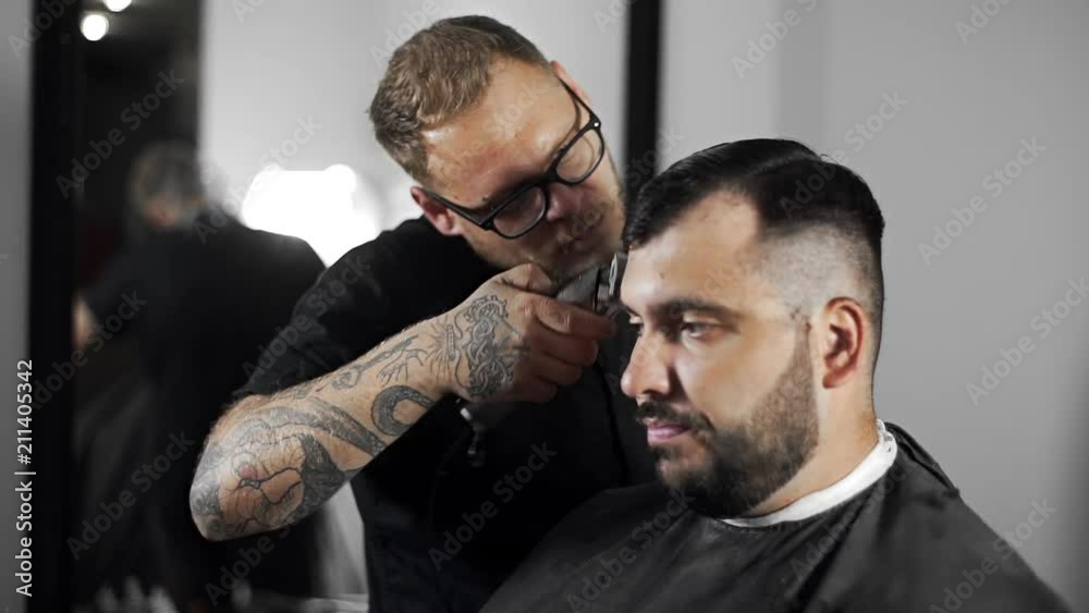 Tattoed barber makes haircut for customer at the barber shop by using ...