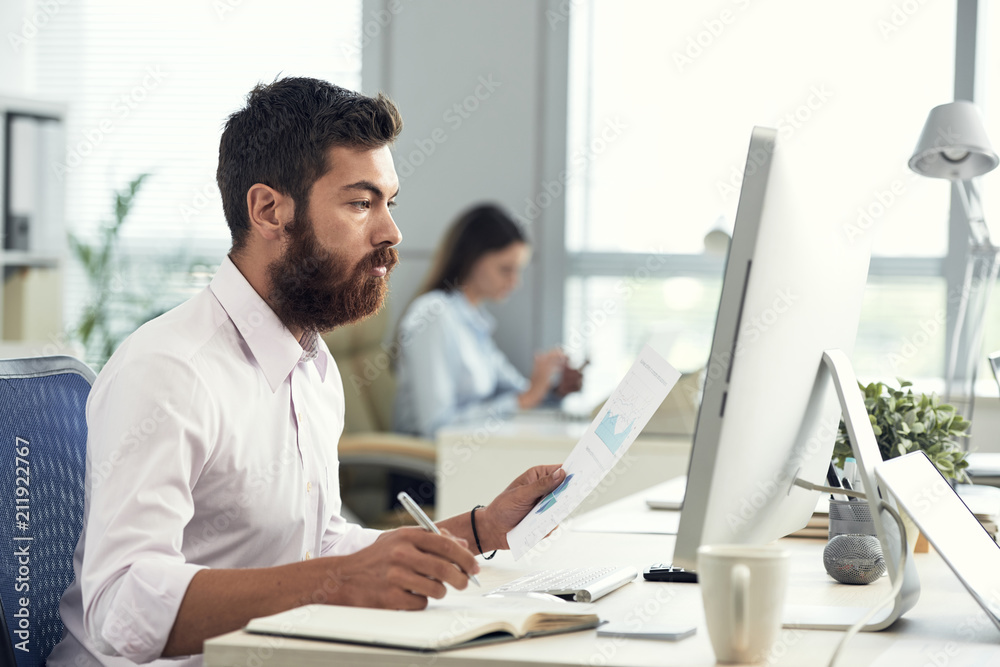 Side view of bearded man watching computer while working with papers in ...