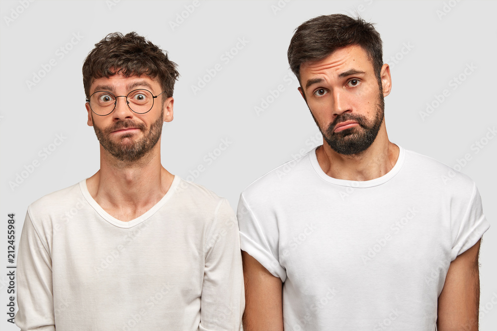 Puzzled sad two bearded young guys stand closely to each other, express ...