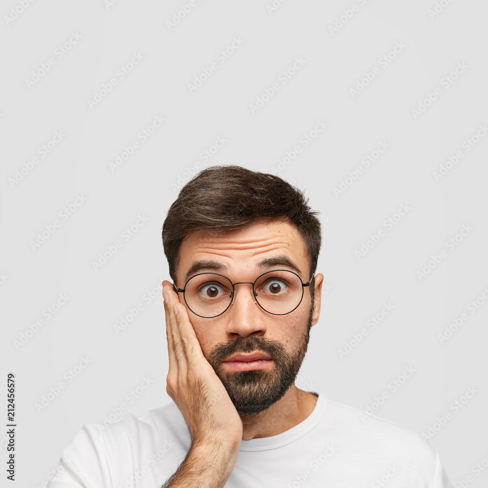 Vertical shot of stupefied Caucasian male touches cheek and stares at ...