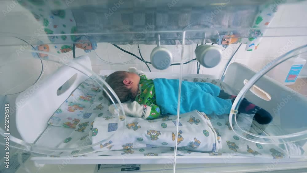 A baby is in incubator, close up. Newborn child in clothes lays in a ...