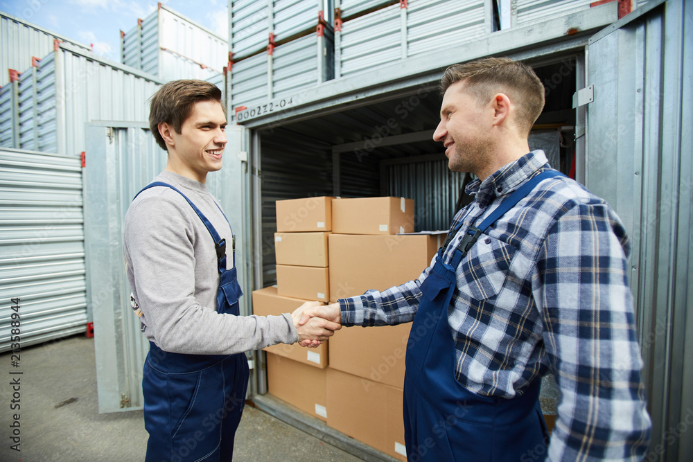 Happy positive handsome cargo coworkers in overalls shaking hands while ...