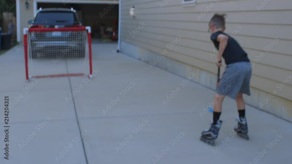 Tracking shot of a boy playing roller hockey in the driveway as he ...