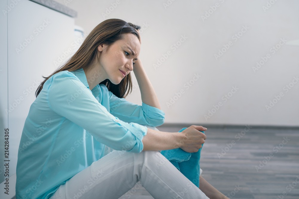 Young woman sitting on kitchen floor holding her head and crying, upset ...