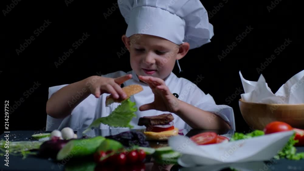 Vidéo Stock Cute boy is eating a burger while cooking. He wears chefs ...