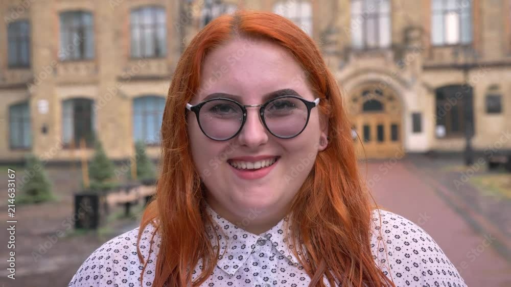 Fat ginger girl with glasses is standing in rainy weather, watching at camera, smiling, building ...