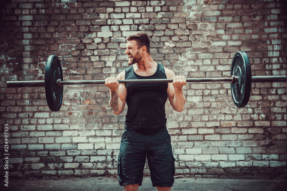 Athletic man working out with a barbell in front of brick wall ...
