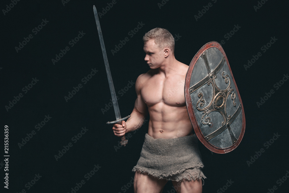 Bodybuilder man posing with a sword and shield isolated on black ...