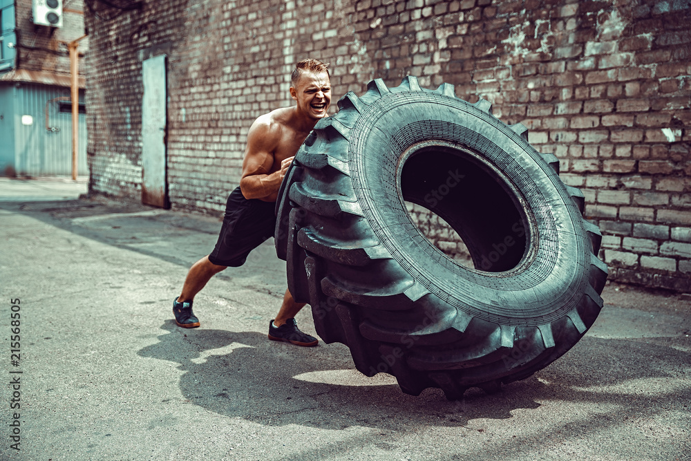 Muscular fitness shirtless man moving large heavy tire in street gym ...