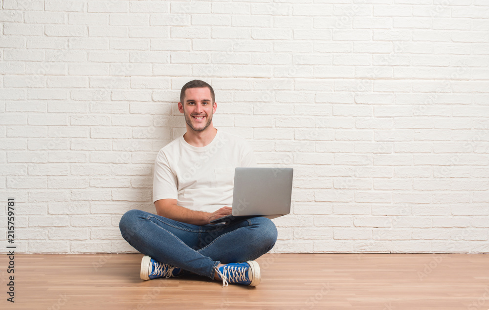 Young caucasian man sitting over white brick wall using computer laptop ...