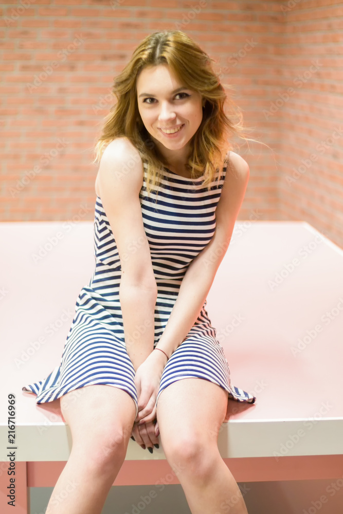 beautiful girl is sitting on the edge of the table in a dress. smiles ...