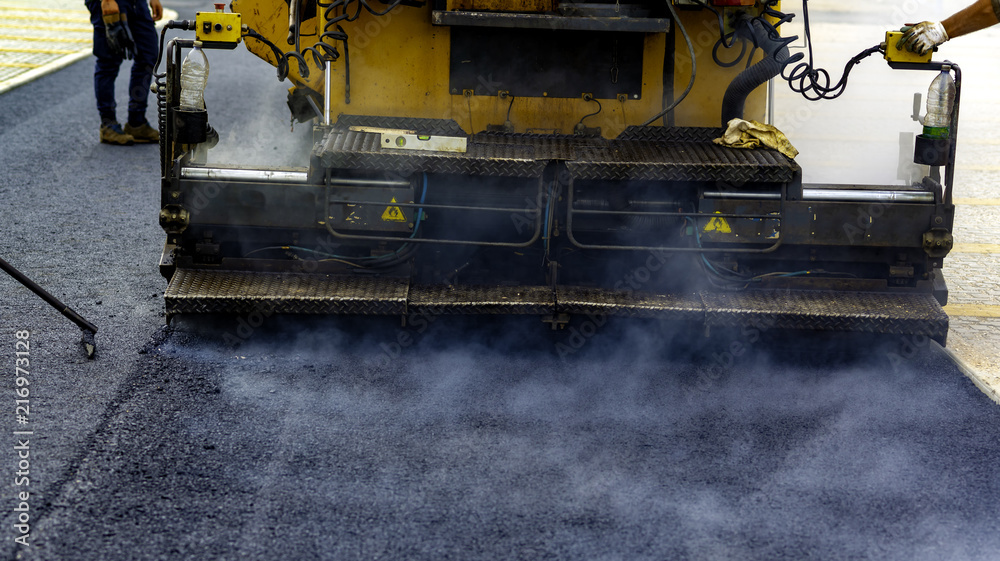 Workers regulate tracked paver laying asphalt heated to temperatures ...