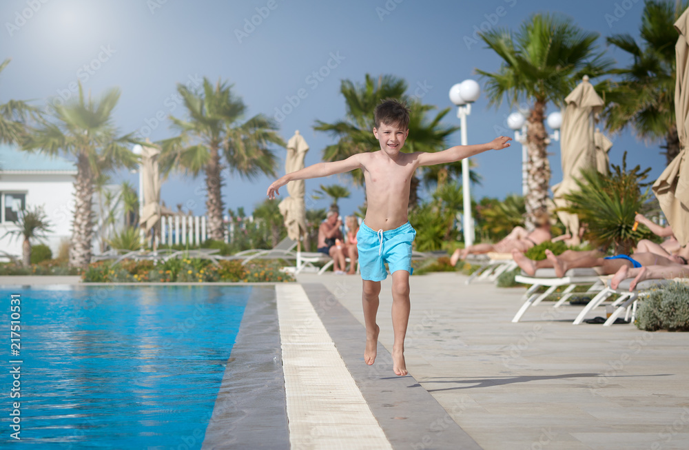 Smiling European boy running along swimming pool at resort in summer ...