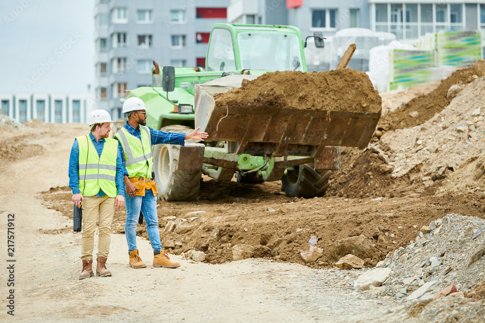 Two men in helmets and waistcoats standing near digger and watching ...
