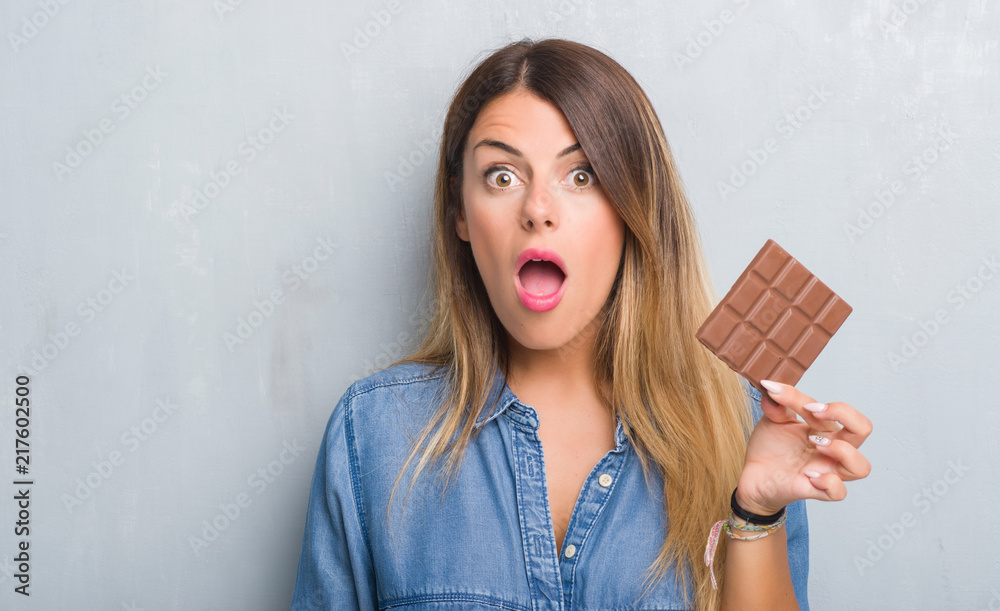 Young adult woman over grey grunge wall eating chocolate bar scared in ...