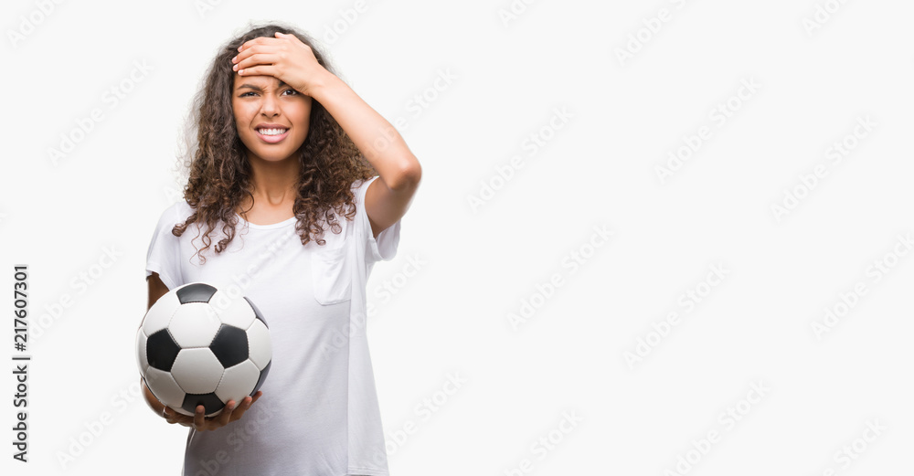 Young hispanic woman holding soccer football ball stressed with hand on ...