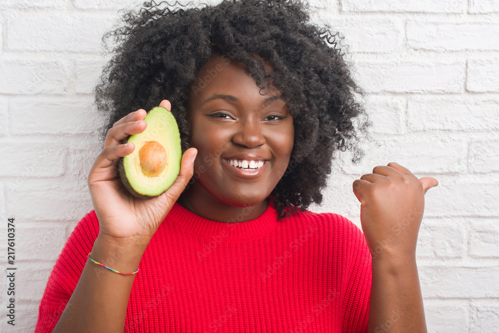 Young african american woman over white brick wall eating avocado ...