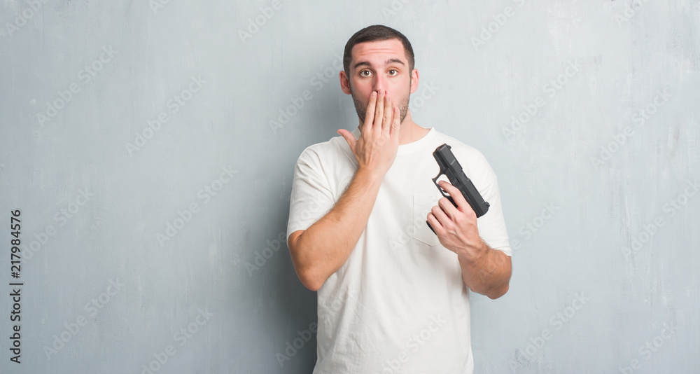 Young caucasian criminal man over grey grunge wall holding gun cover ...