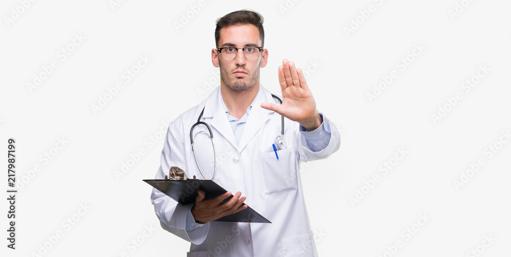Handsome young doctor man holding a clipboard with open hand doing stop ...