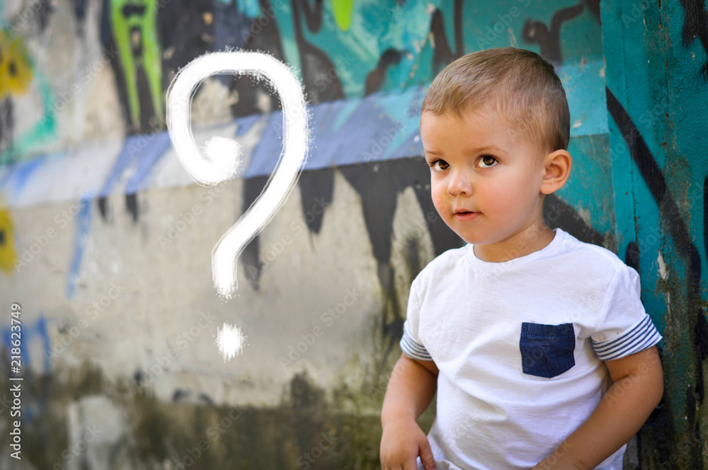 A two-year-old baby boy in hip hop style clothes sits under a wall with ...