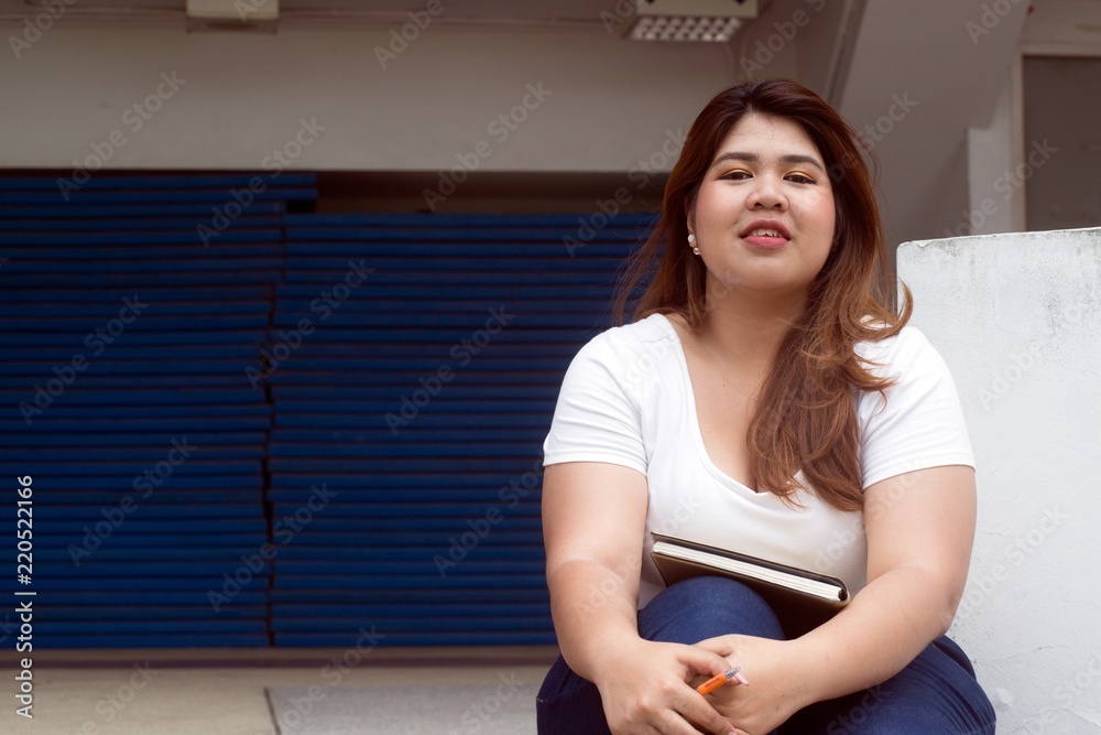 Portrait of Pretty Asian smiley fat woman sit and thinking at staircase ...