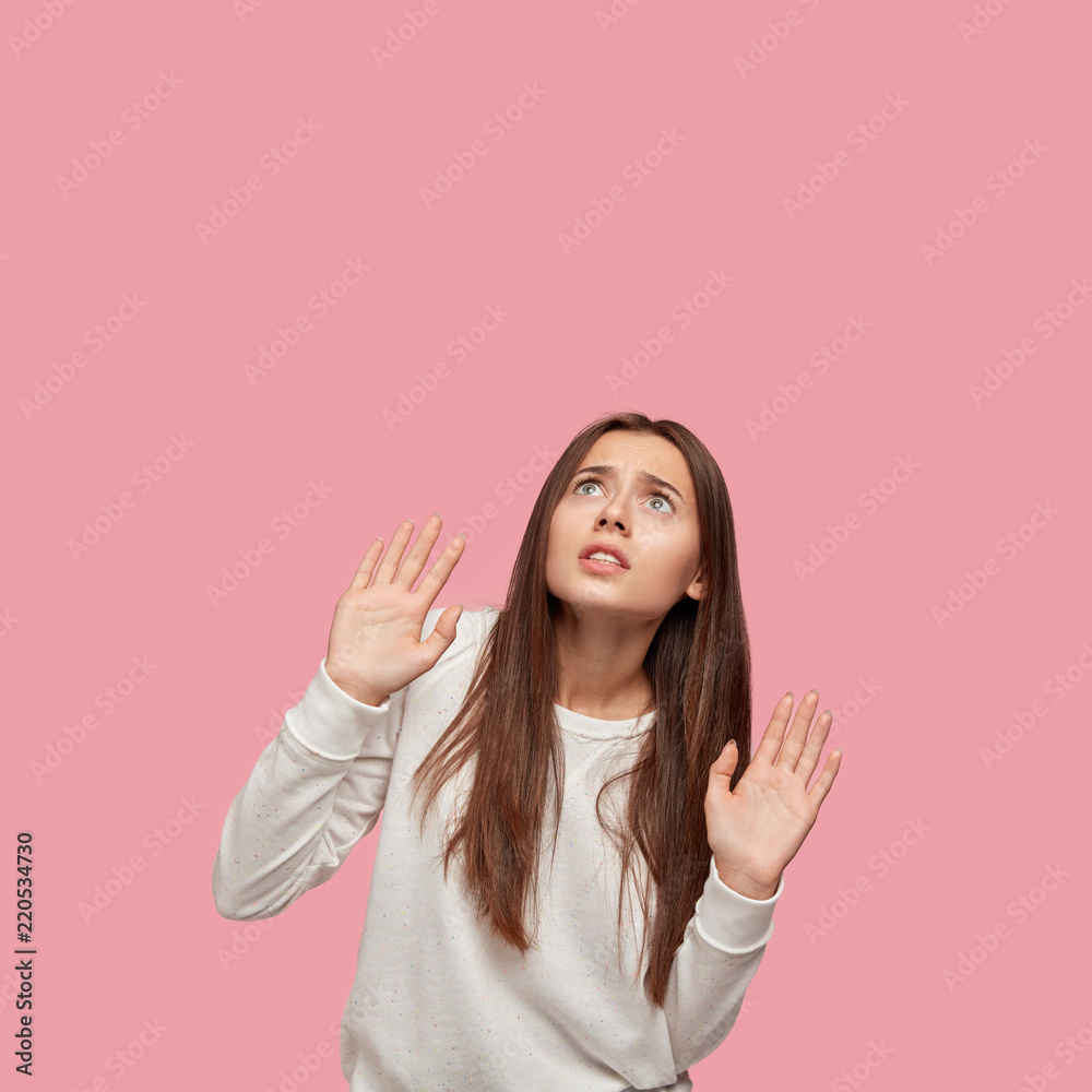 Frightened perplexed embarrassed young girl looks upwards, keeps palms ...
