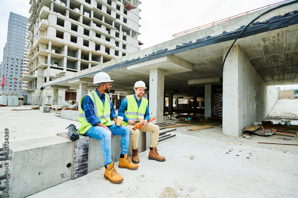 Positive handsome young builders in safety vests and hardhats sitting ...