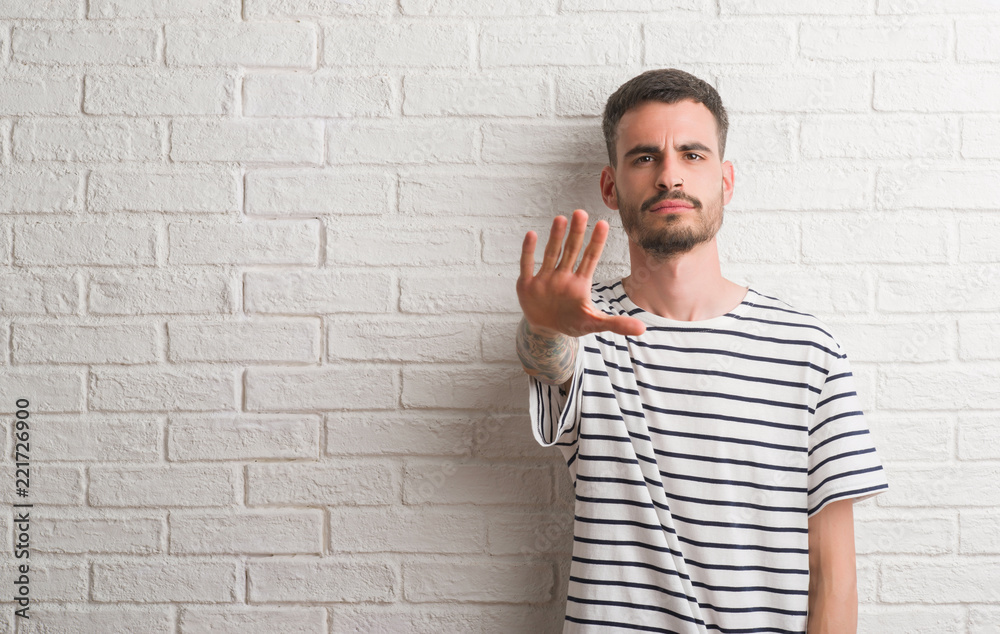Young adult man standing over white brick wall with open hand doing ...