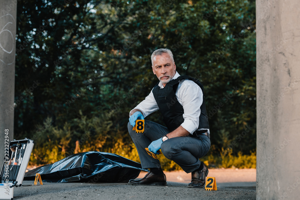 serious male police officer in latex gloves sitting at crime scene with ...