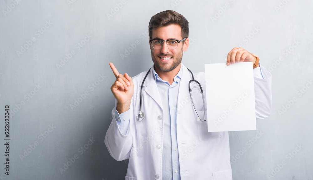 Handsome young doctor man over grey grunge wall holding blank sheet ...