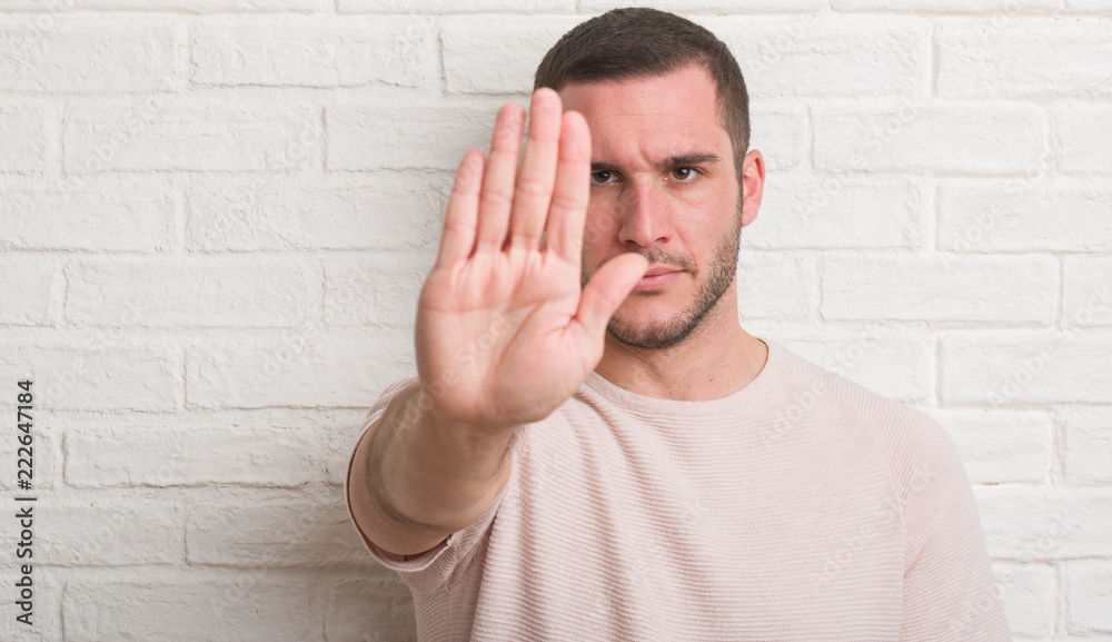 Young caucasian man standing over white brick wall with open hand doing ...