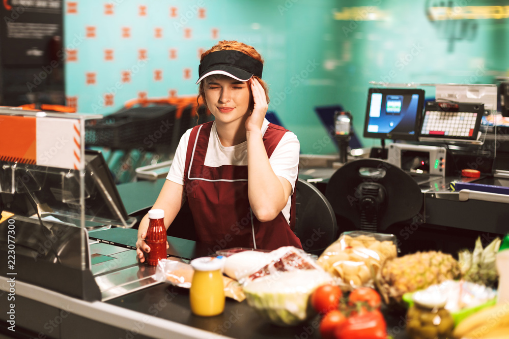Young female cashier in uniform with headache working in supermarket ...