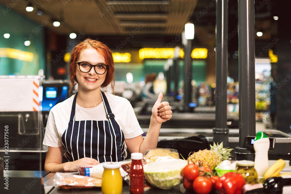 Young smiling female cashier in eyeglasses and striped apron with ...
