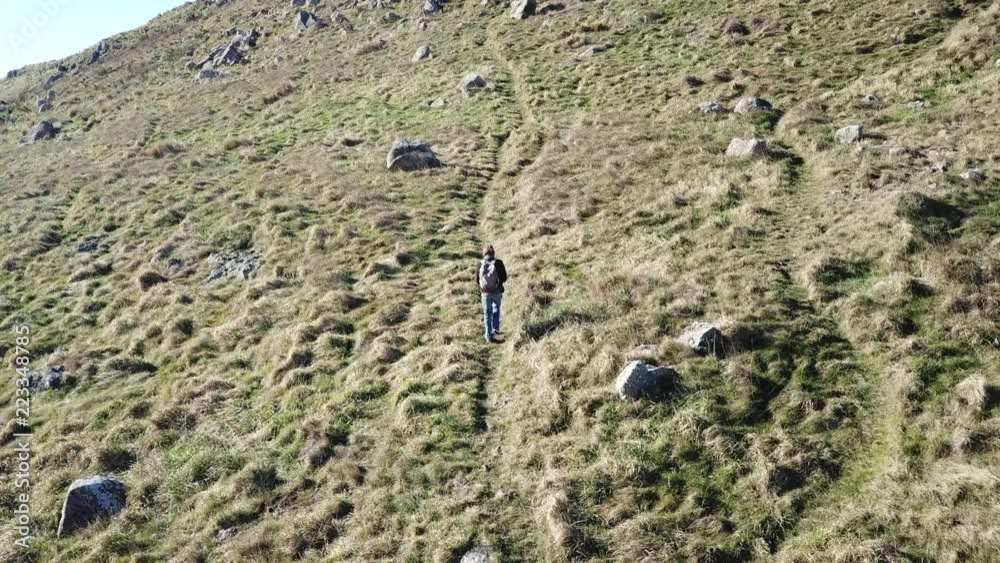 Drone footage above man walking on a track up a steep hill to a rocky ...