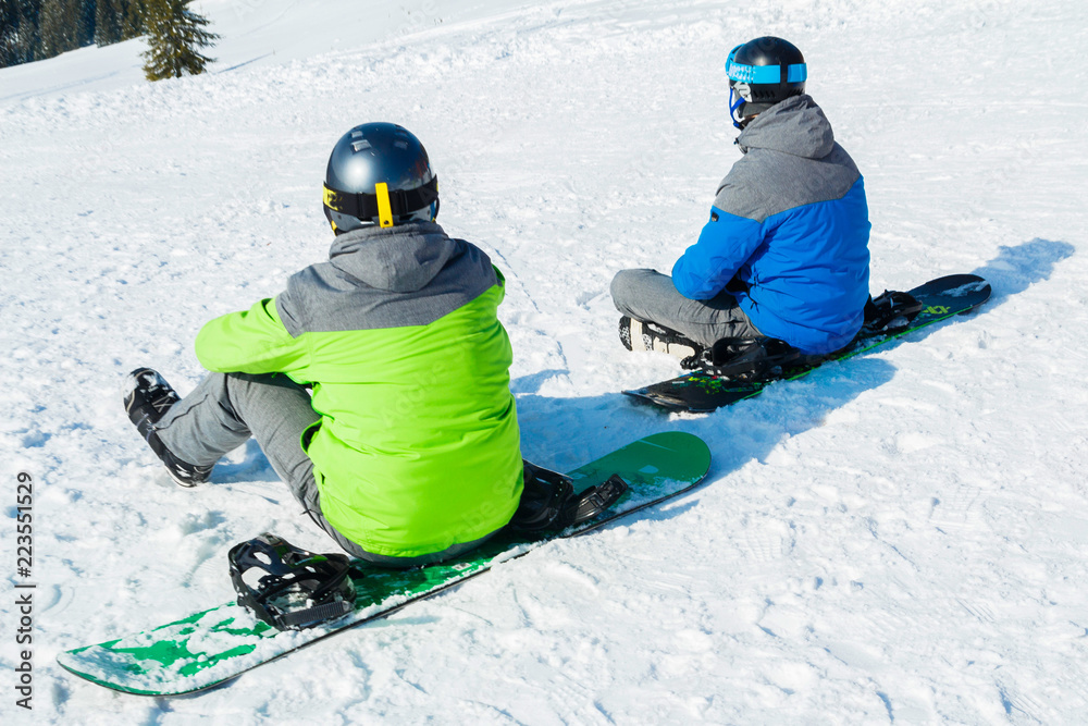 Two snowboarders are sitting on the snow at the top of the mountain and ...