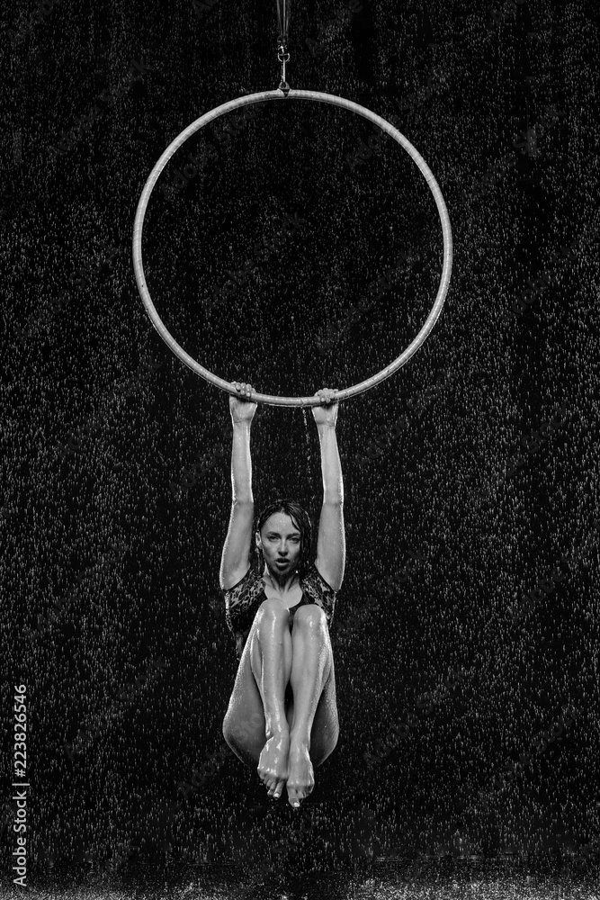 Beautiful female acrobat hanging on aerial hoop under rain on black ...